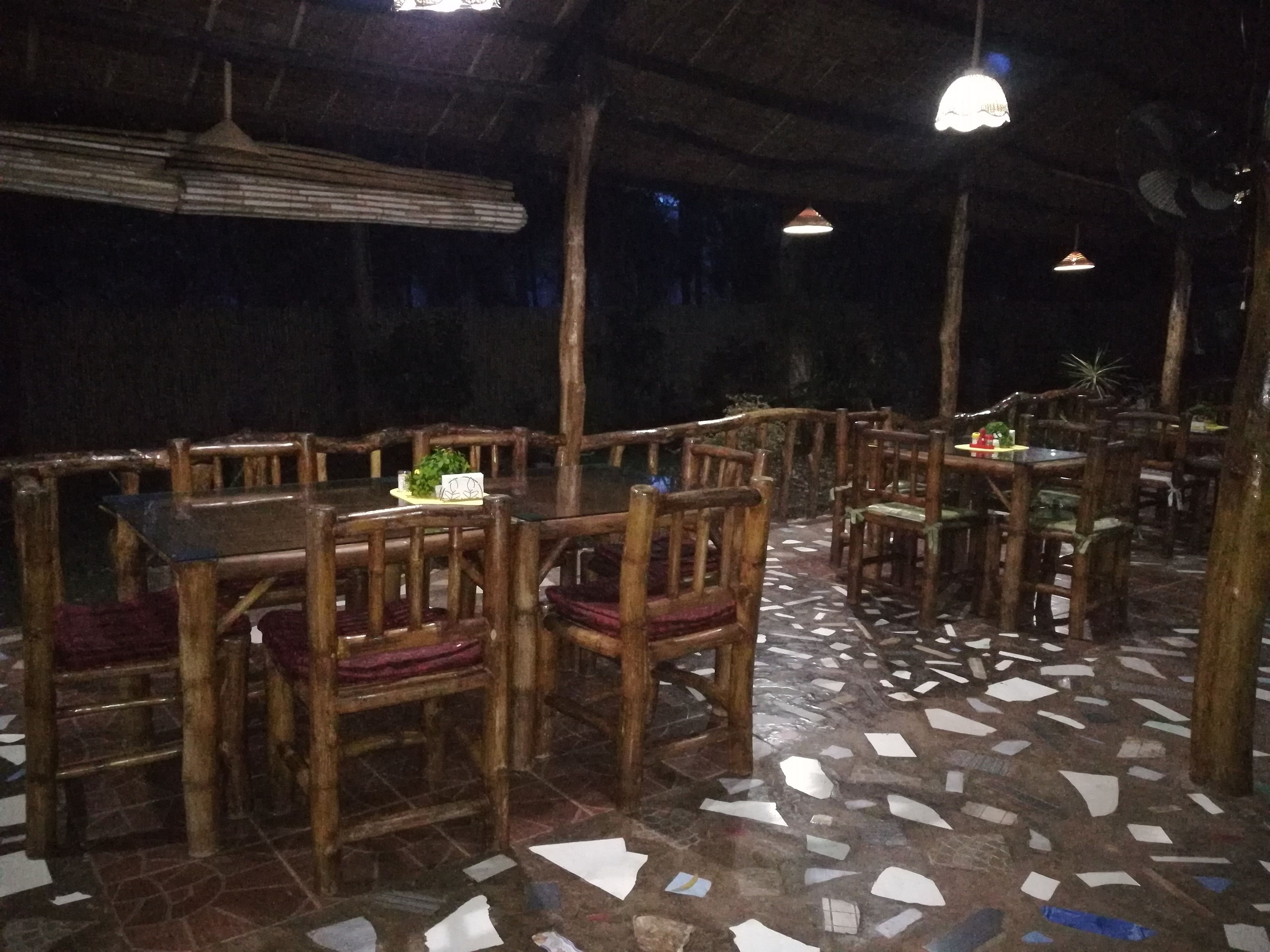 Um restaurante com mesas e cadeiras de madeira, piso ladrilhado e luminárias pendentes.