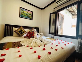 Cama bem arrumada com um cisne decorativo e pétalas de rosa, um quadro de paisagem emoldurado e uma janela com portadas.