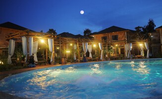 Uma área de piscina à noite, com vista clara para a lua, pérgulas de madeira com cortinas esvoaçantes e uma piscina refletindo o céu.