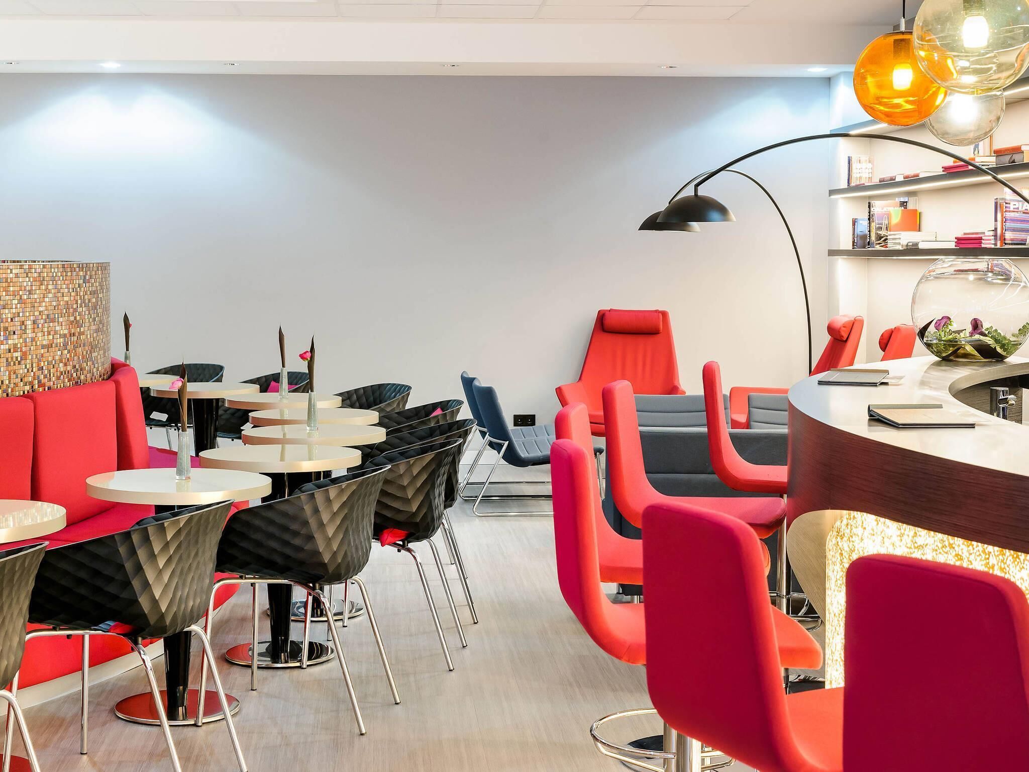 Uma área de estar moderna com poltronas estofadas em vermelho e preto, mesas redondas e uma estante de livros com livros.