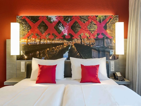 Quarto de hotel com uma cama grande, duas almofadas vermelhas, uma mesa de cabeceira com telefone e uma televisão fixada na parede exibindo uma vista urbana.