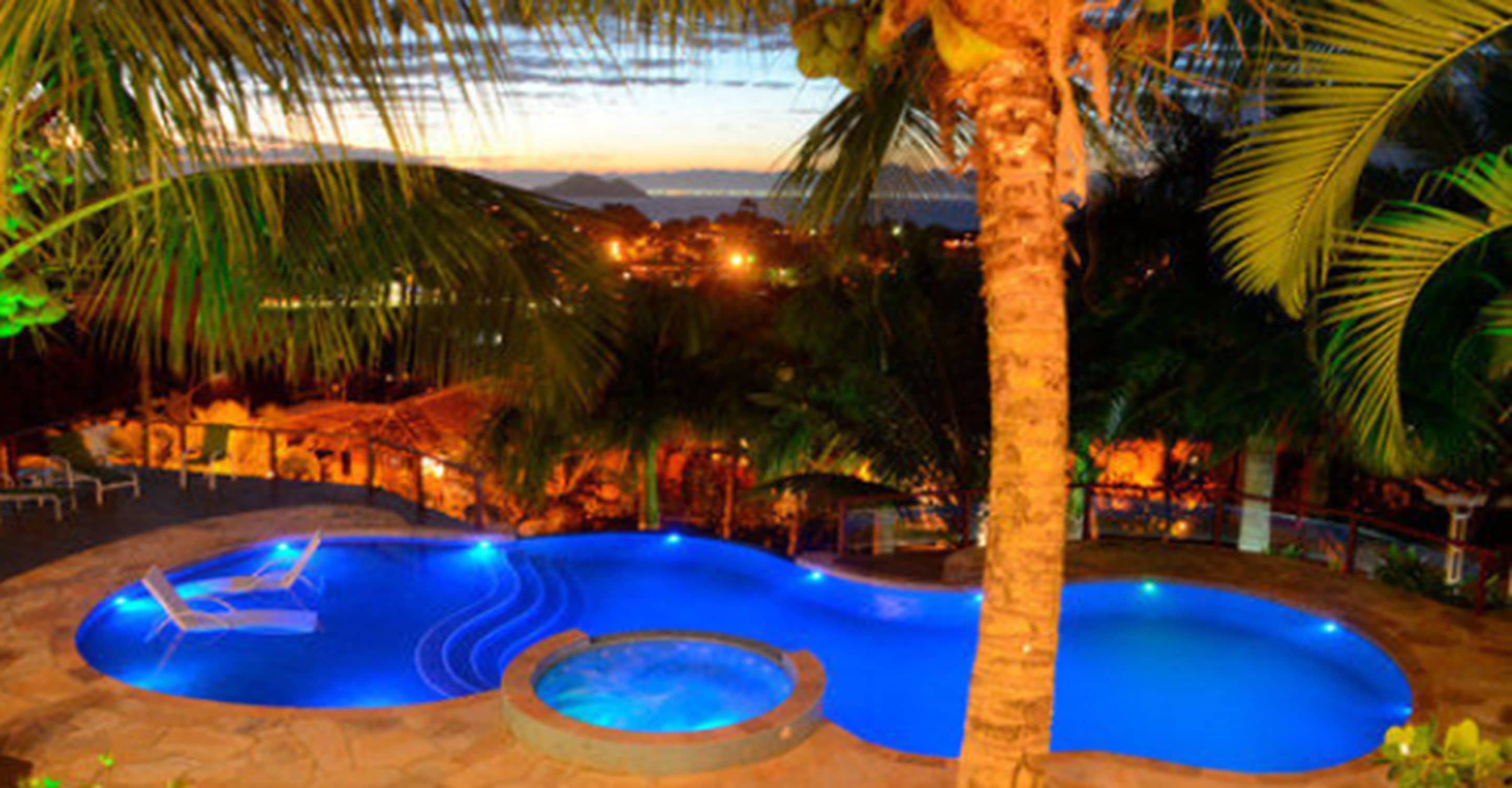 Piscina com hidromassagem, rodeada por palmeiras e vista para o horizonte urbano ao entardecer.