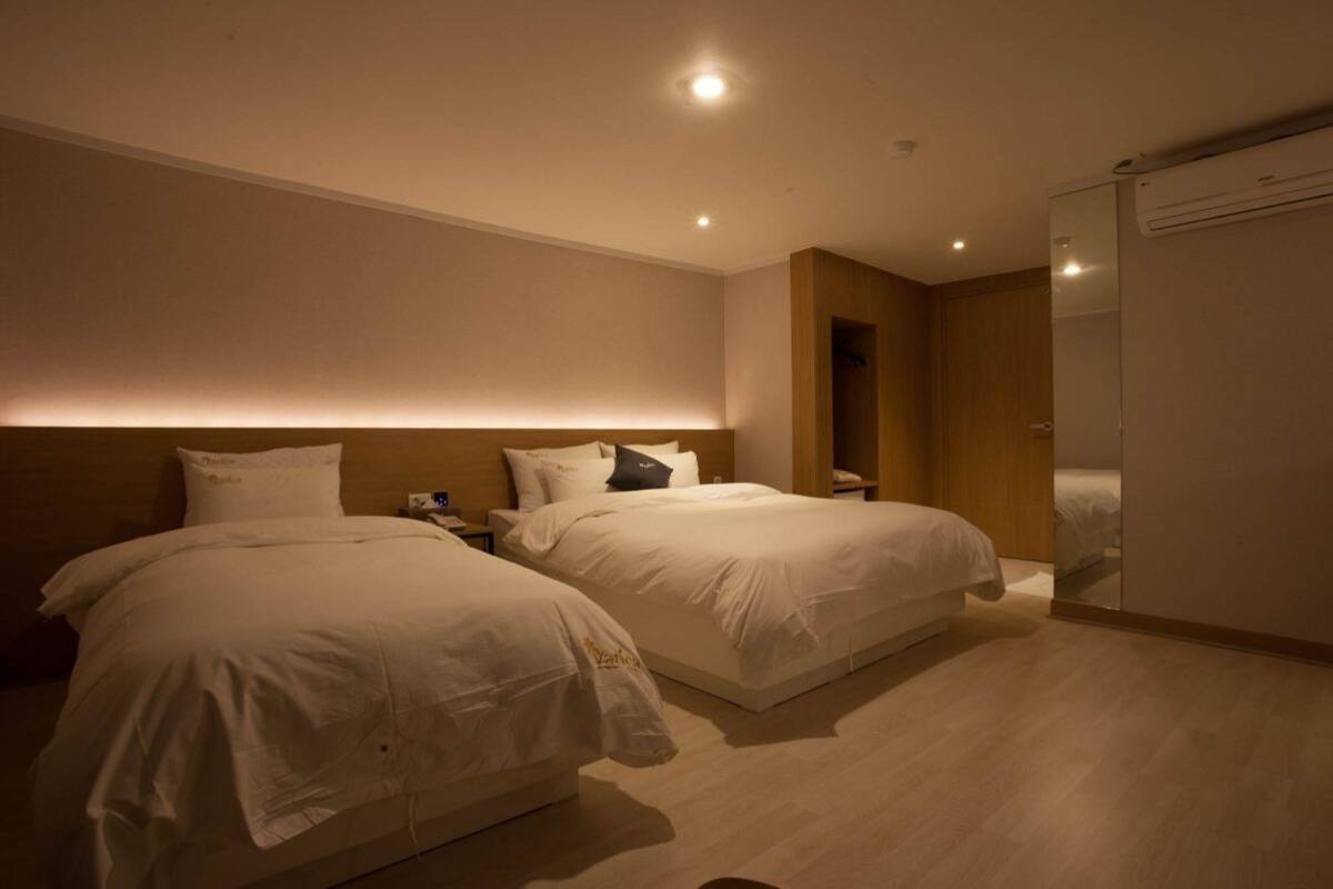 Quarto de hotel com duas camas, cabeceira de madeira, guarda-roupa e mesa de cabeceira com abajur.
