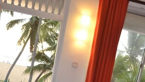 Quarto com cama, janela com cortina, varanda com guarda-corpo e vista para palmeiras e praia.