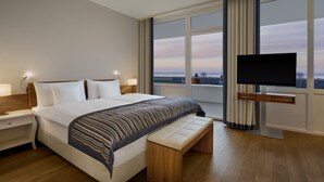 Quarto de hotel moderno com uma cama grande, uma televisão de tela plana na parede, uma mesa de cabeceira com abajur e vista da cidade através de grandes janelas.