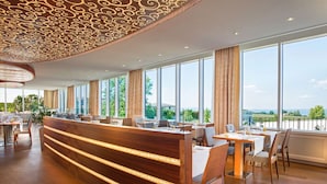 Um hall de hotel espaçoso com um balcão de recepção de madeira, grandes janelas com vista para o exterior e um teto alto com padrões elaborados.