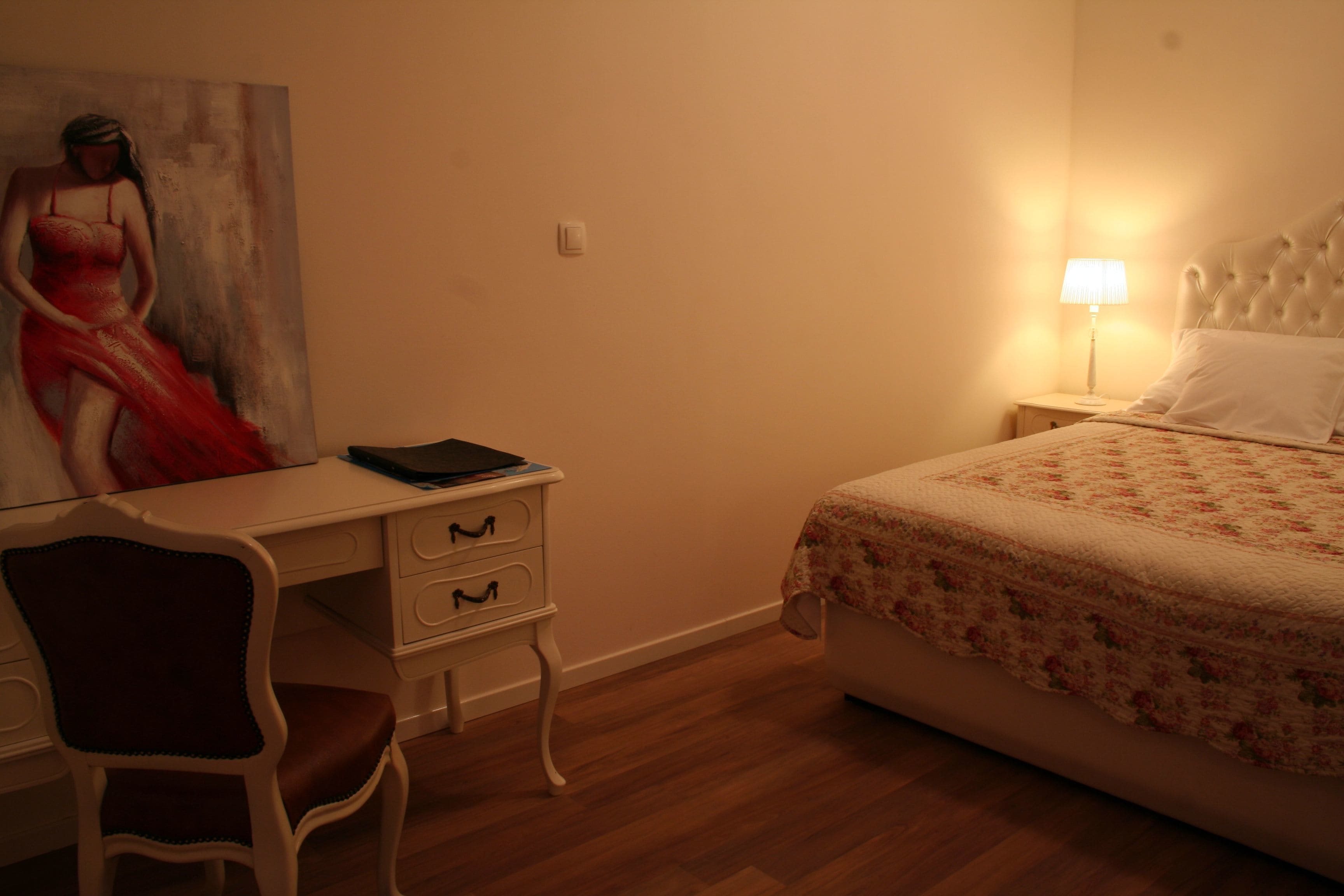 Quarto com cama, escrivaninha, cadeira, mesa de cabeceira com abajur e um quadro na parede.