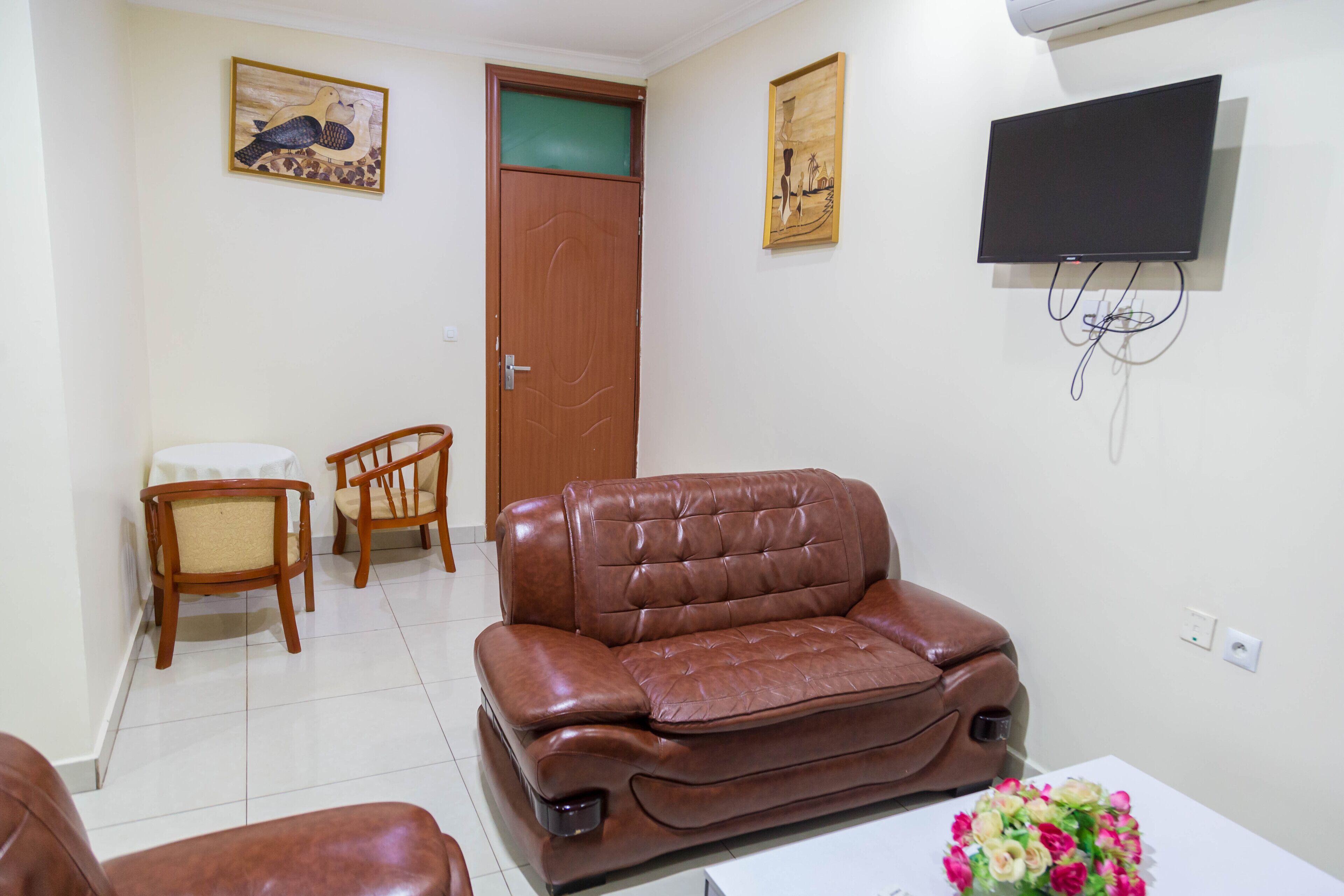 Uma sala de estar com um sofá de couro marrom, duas cadeiras de madeira, uma mesa pequena e uma TV fixada na parede.