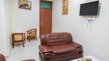 Uma sala de estar com um sofá de couro marrom, duas cadeiras de madeira, uma mesa pequena e uma TV fixada na parede.