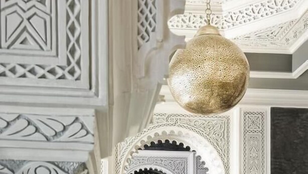 Um corredor ricamente decorado, com arcos brancos detalhados e um lustre esférico pendurado.