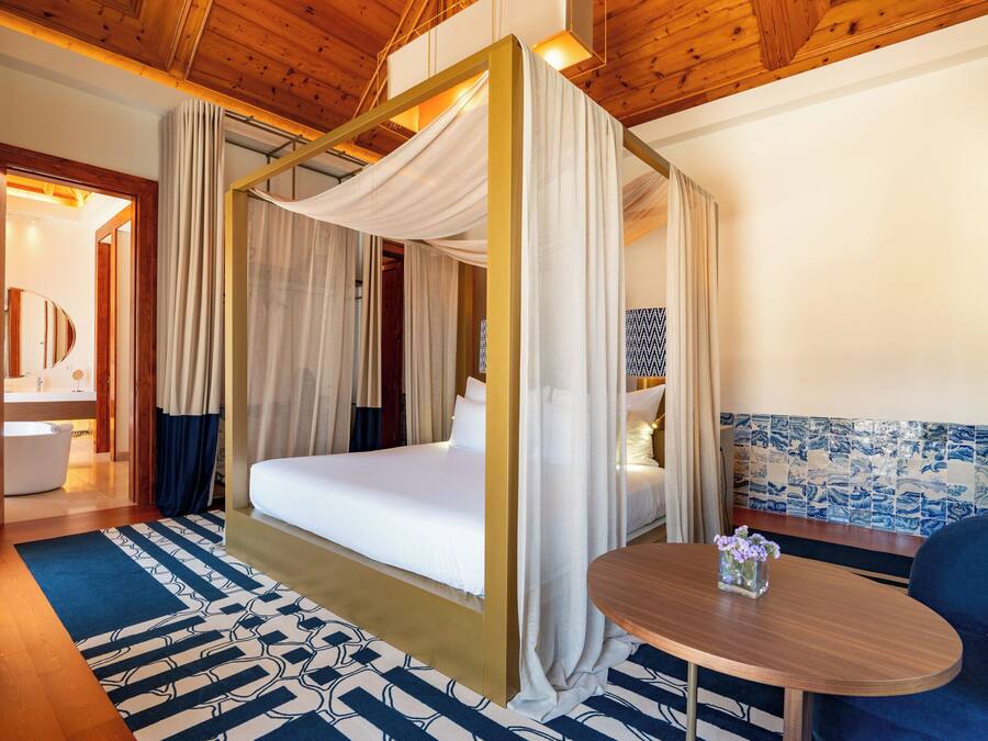 Uma cama de dossel com lençóis brancos, uma cabeceira de madeira e um tapete azul e branco estampado. Uma mesa redonda de madeira com um vaso de flores está visível.