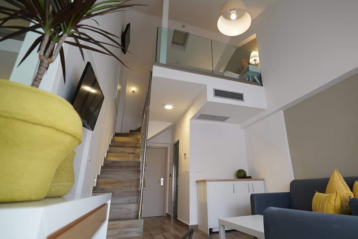 Sala moderna com escada para um andar superior, sofá com almofadas amarelas e um vaso com planta.