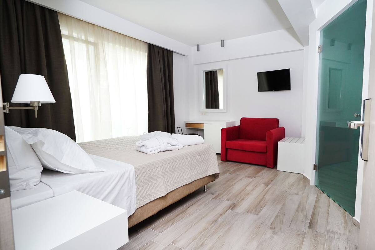 Quarto de hotel com cama, uma cadeira vermelha, escrivaninha, luminária e televisão.