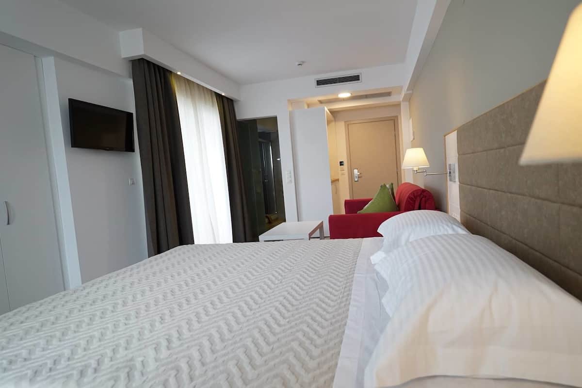 Quarto de hotel com uma cama grande, uma poltrona vermelha, uma mesa pequena e uma televisão na parede.