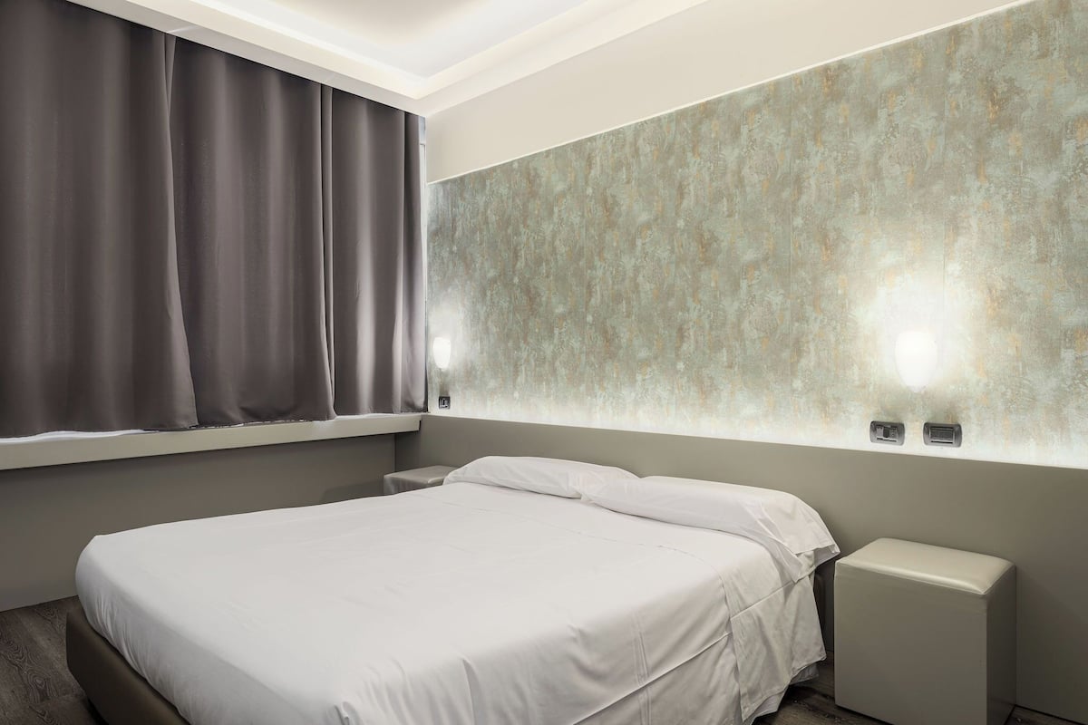 Quarto de hotel moderno com uma cama grande, janela com cortinas, parede texturizada e uma pequena mesa de cabeceira.