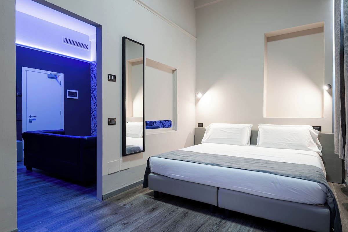 Quarto de hotel moderno com uma cama grande, uma mesinha de cabeceira, um espelho e uma porta para um banheiro.