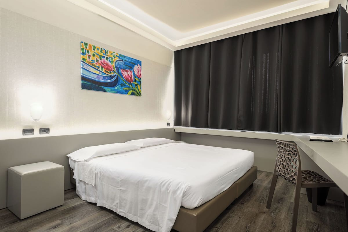 Quarto de hotel com cama, mesa de cabeceira pequena, cadeira, escrivaninha, um quadro na parede e janela com cortinas.