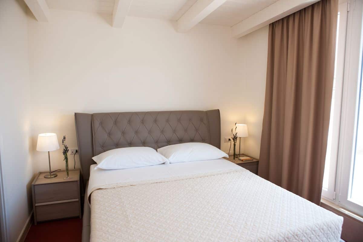 Cama bem arrumada com cabeceira estofada, duas mesas de cabeceira com abajures e uma janela com cortinas.
