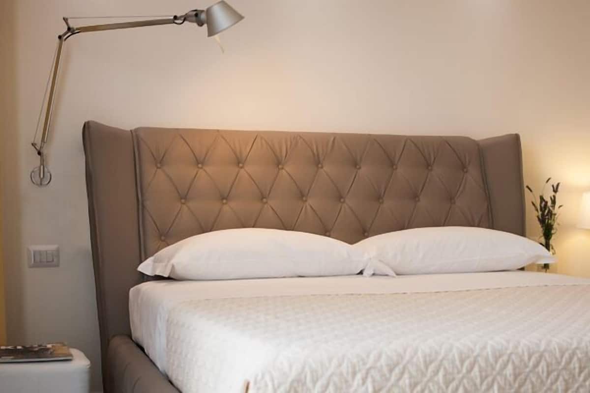 Cama bem arrumada com cabeceira estofada, luminária de cabeceira e mesinha de apoio com uma revista.