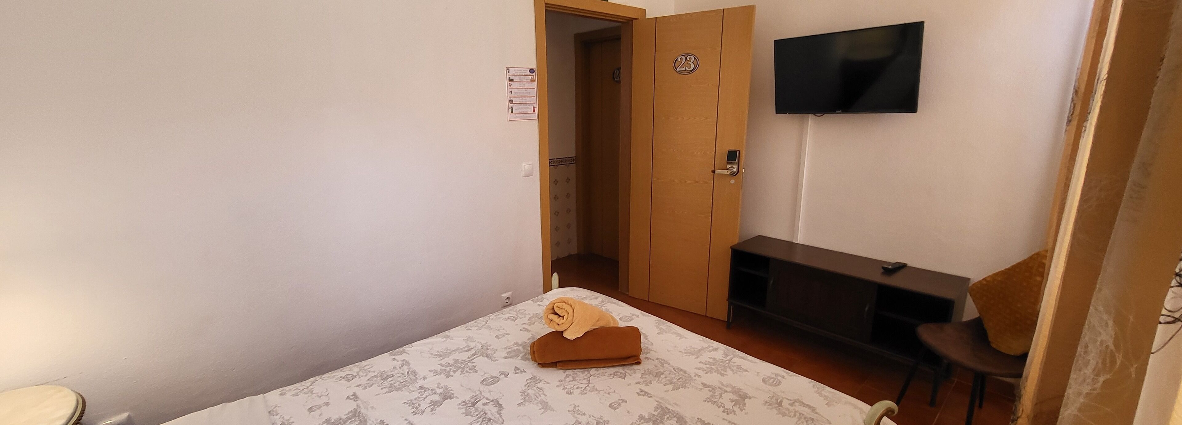 Quarto com cama, televisão presa na parede, porta de madeira e pequena escrivaninha.