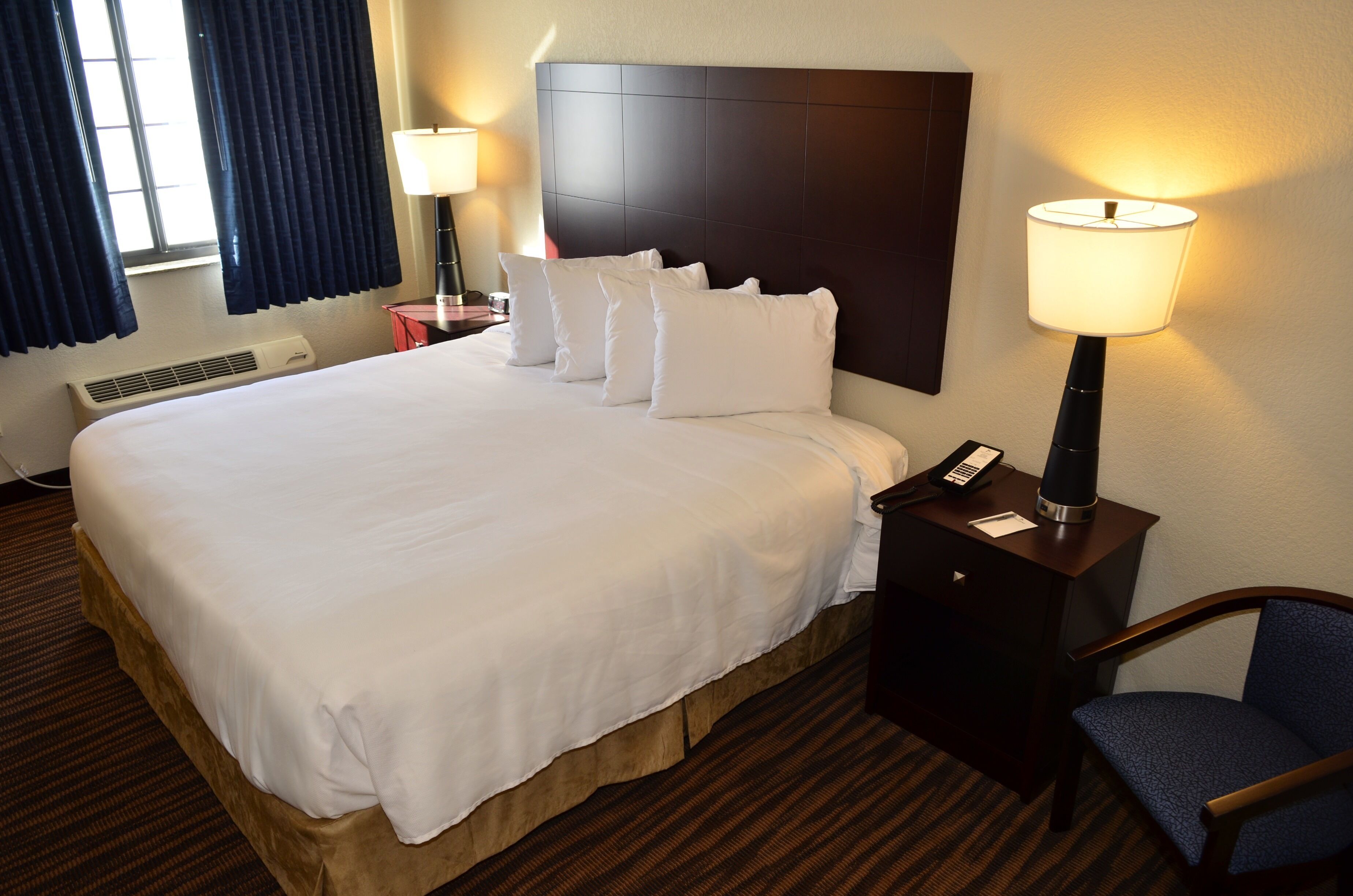 Quarto de hotel com uma cama grande, abajur, cadeira e mesa com telefone e cafeteira.