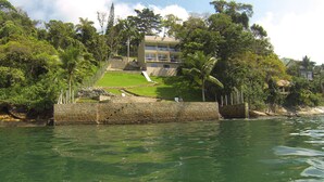 Um imóvel à beira-mar com muro de pedra, vegetação exuberante e um edifício de vários andares com varandas.