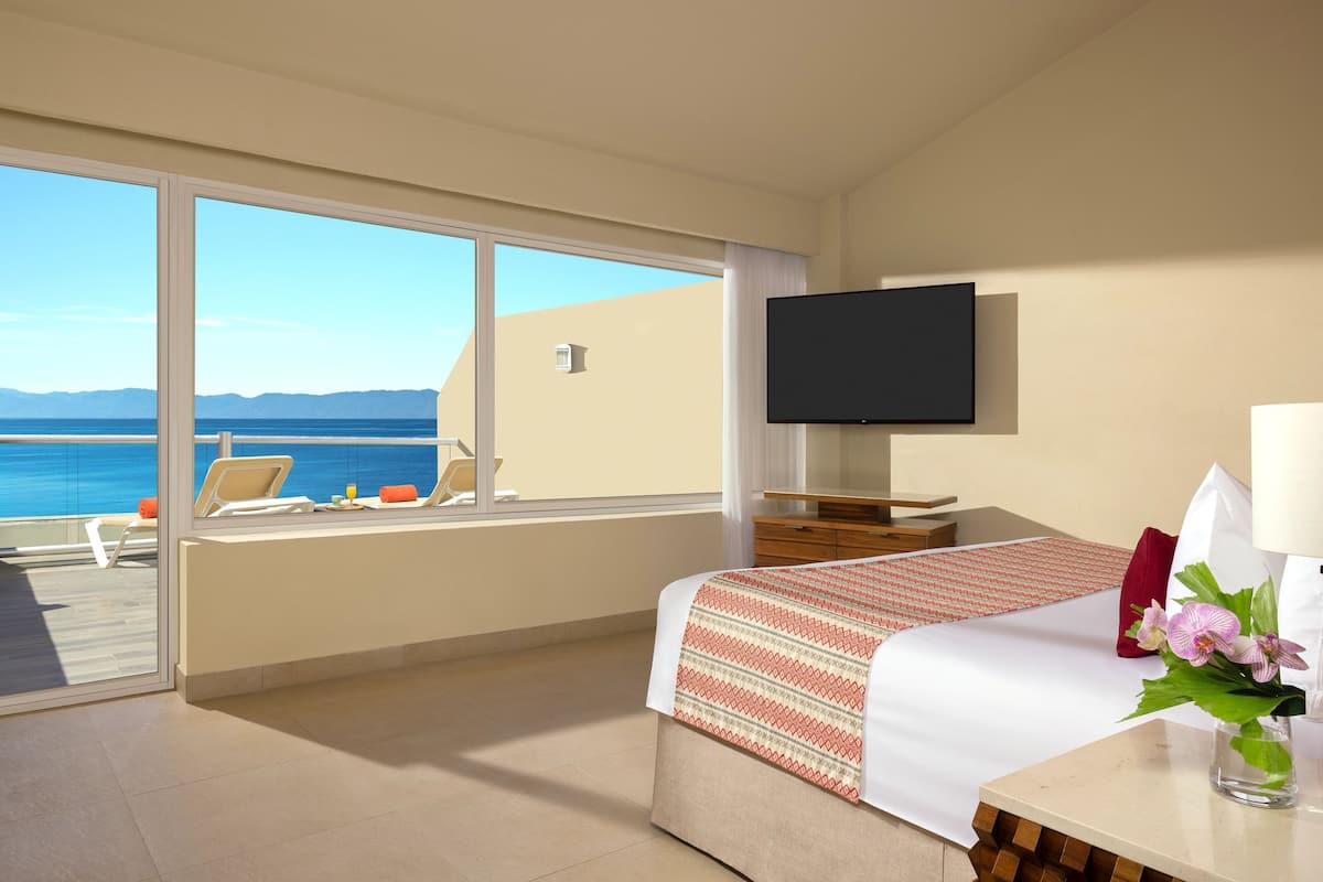 Quarto de hotel com uma cama, televisão, escrivaninha com luminária e vista para o mar.