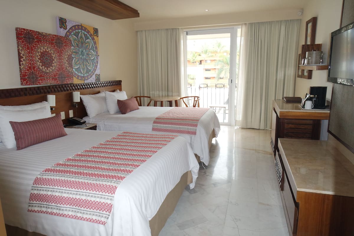 Quarto de hotel com duas camas, uma mesa de escritório com televisão e varanda com cortinas.