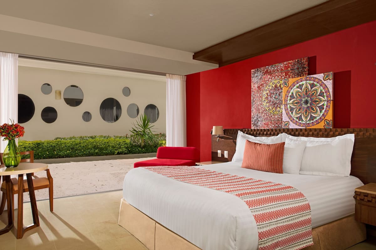 Quarto de hotel moderno com uma cama grande, detalhes em vermelho, vista para um jardim ajardinado e uma peça de arte única na parede.