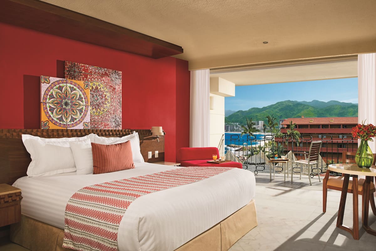 Quarto de hotel com uma cama grande, uma varanda com vista para as montanhas e uma área de refeições com mesa e cadeiras.