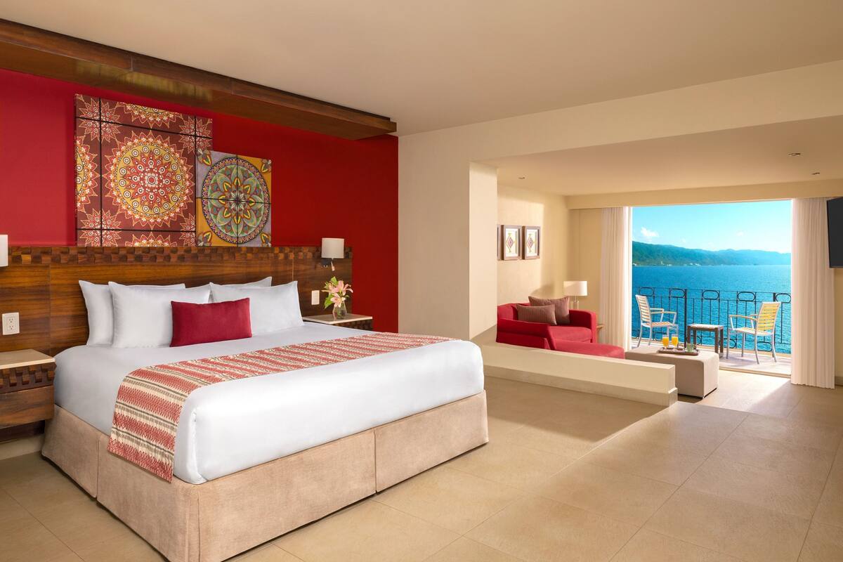 Quarto de hotel com cama grande, parede de destaque vermelha, varanda com vista para o mar e cabeceira de madeira.