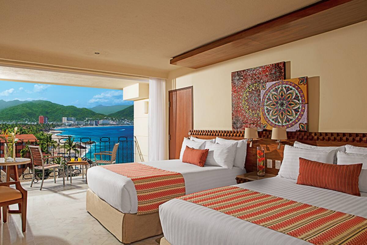 Quarto de hotel com duas camas, vista para o oceano e área de refeições com mesas ao ar livre.