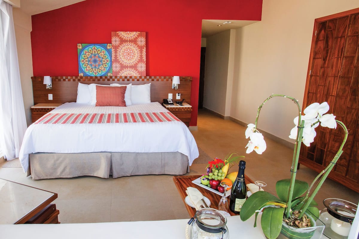 Quarto de hotel com cama grande, cabeceira de madeira, parede em vermelho, um ornamento de parede, mesa de cabeceira com abajur, mesinha de apoio com telefone, pequena mesa com frutas, garrafa de vinho e uma orquídea em vaso.