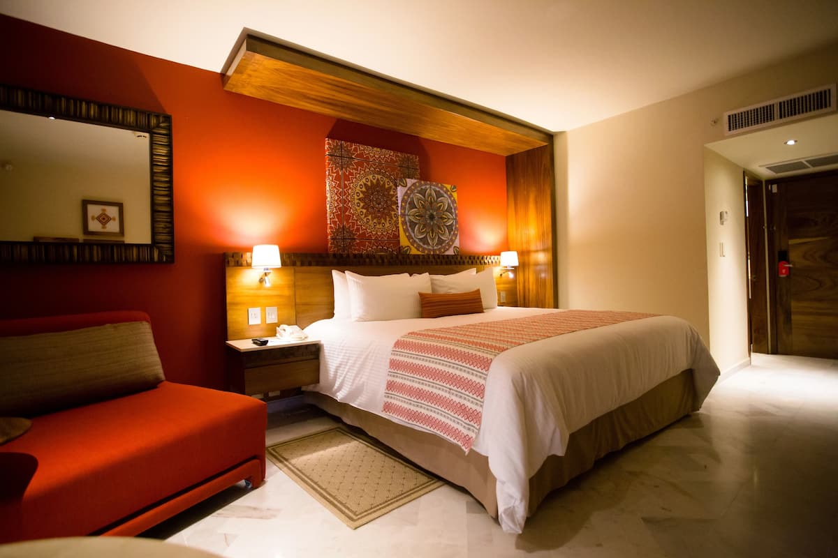Quarto de hotel com cama grande, sofá vermelho, cabeceira de madeira e um objeto decorativo na parede.