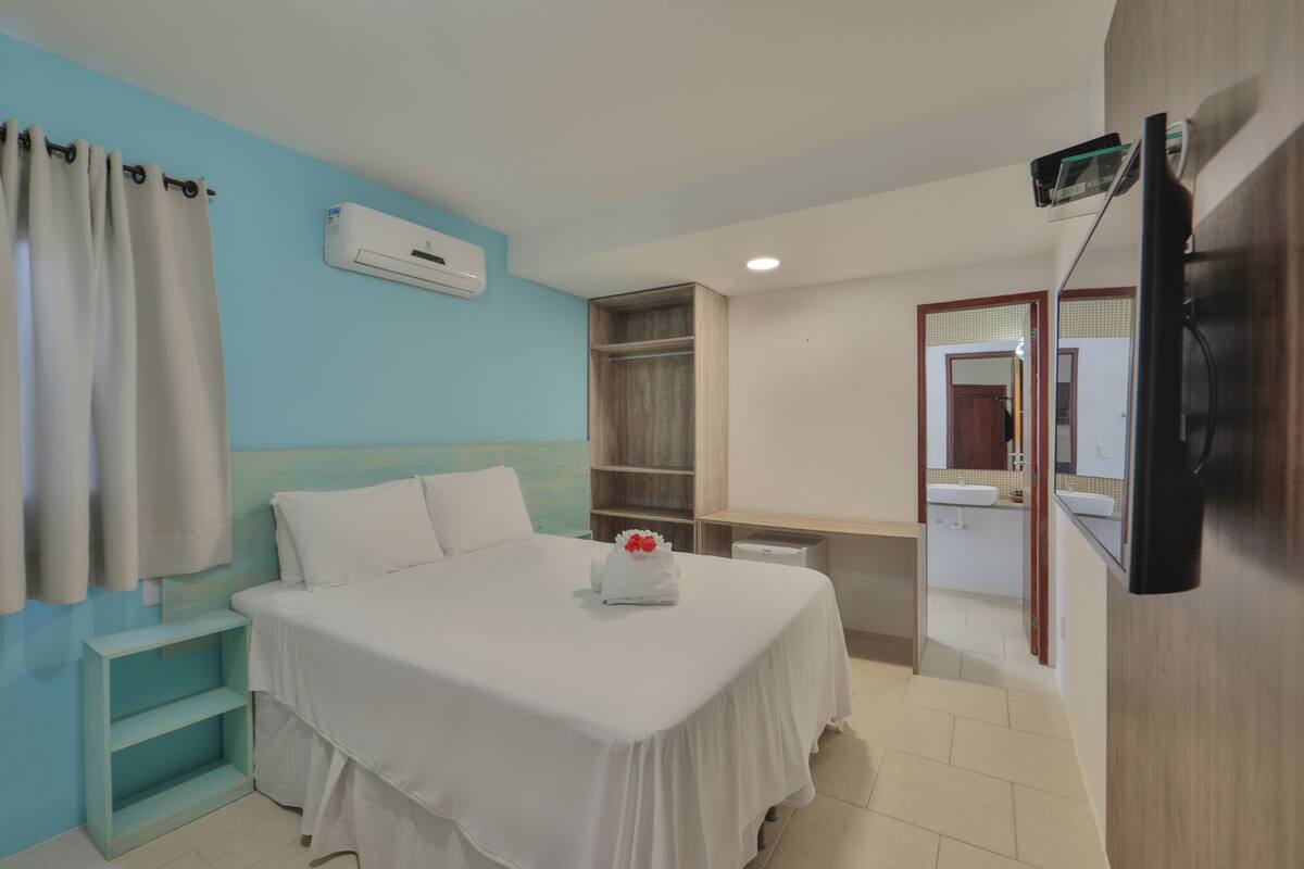 Quarto de hotel com cama, guarda-roupa, secretária e casa de banho visível por uma porta aberta.
