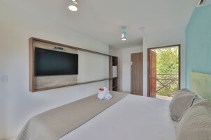 Quarto de hotel com cama, televisão na parede, porta de madeira e vista para o verde lá fora.