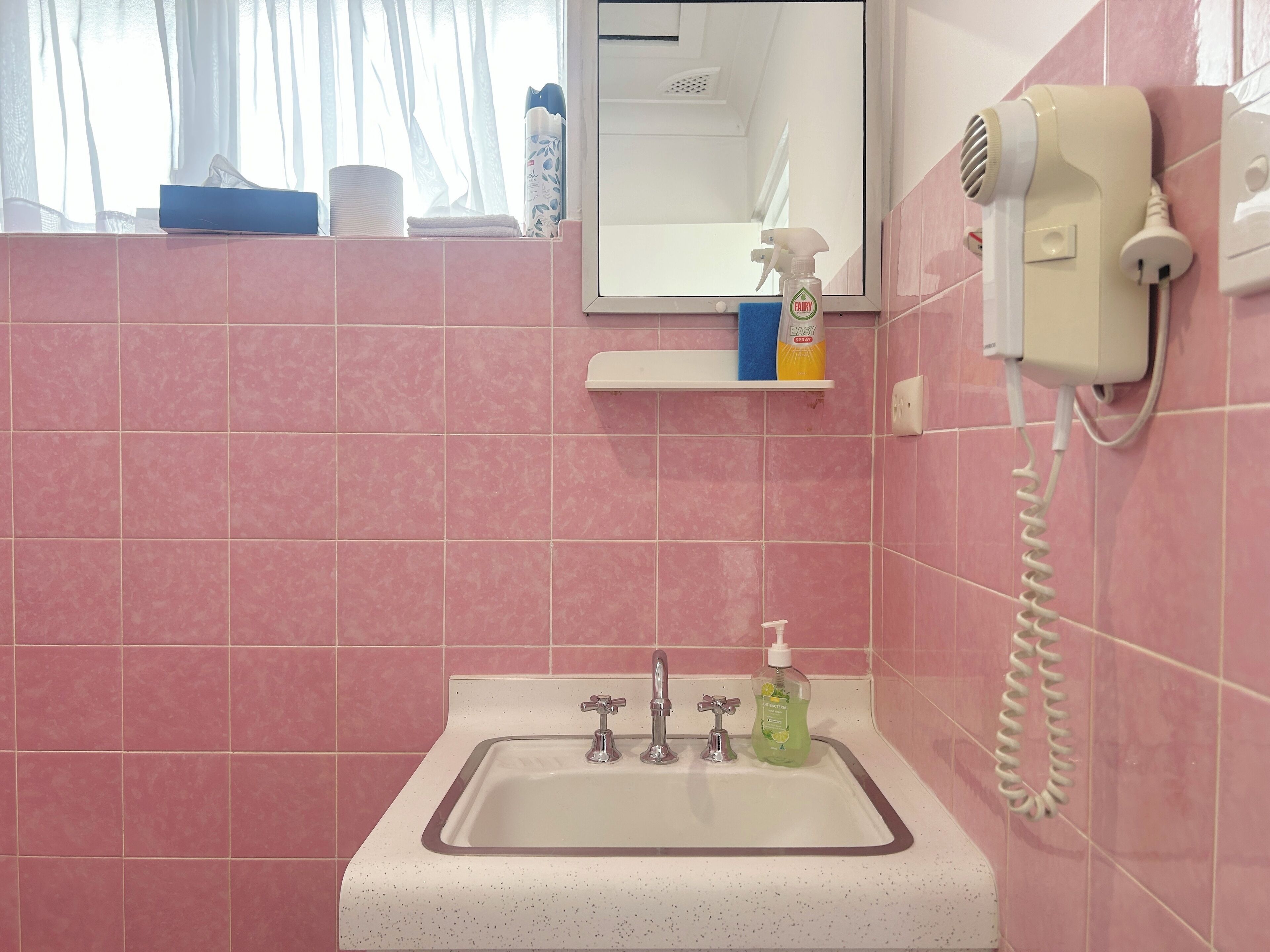 Um banheiro com azulejos cor-de-rosa, uma pia com duas torneiras, um espelho, um secador de cabelo e uma prateleira com produtos de higiene pessoal.