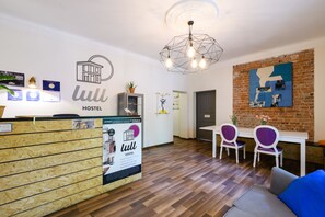 Uma área de receção de hostel com um balcão de madeira, um candeeiro moderno, uma parede de tijolo com obras de arte e um espaço de estar com cadeiras roxas.