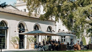 Uma área de refeições ao ar livre com mesas e guarda-sóis, rodeada por um edifício com janelas em arco e telhado em azulejos.