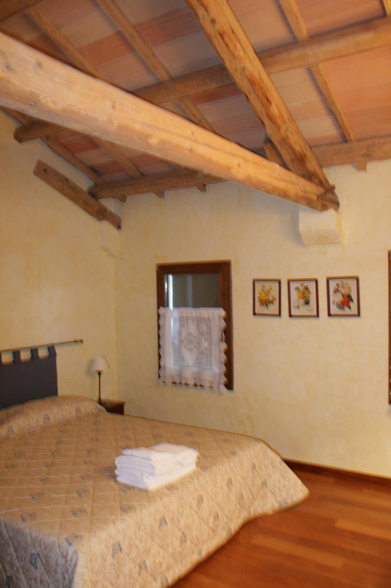 Quarto com teto de madeira, cama com colcha, janela com cortina e três quadros na parede.