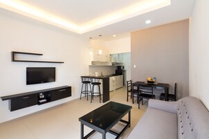 Uma sala de estar moderna com televisão de tela plana, mesa de centro de vidro e sofá. Há também uma cozinha com mesa de jantar e banquetas.