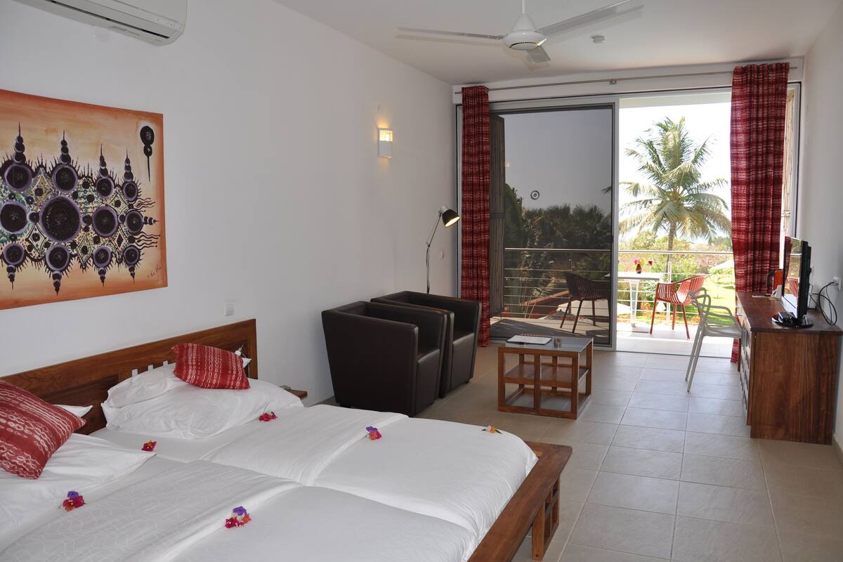 Quarto de hotel com cama, escrivaninha, poltrona, sofá e varanda com vista para palmeiras.