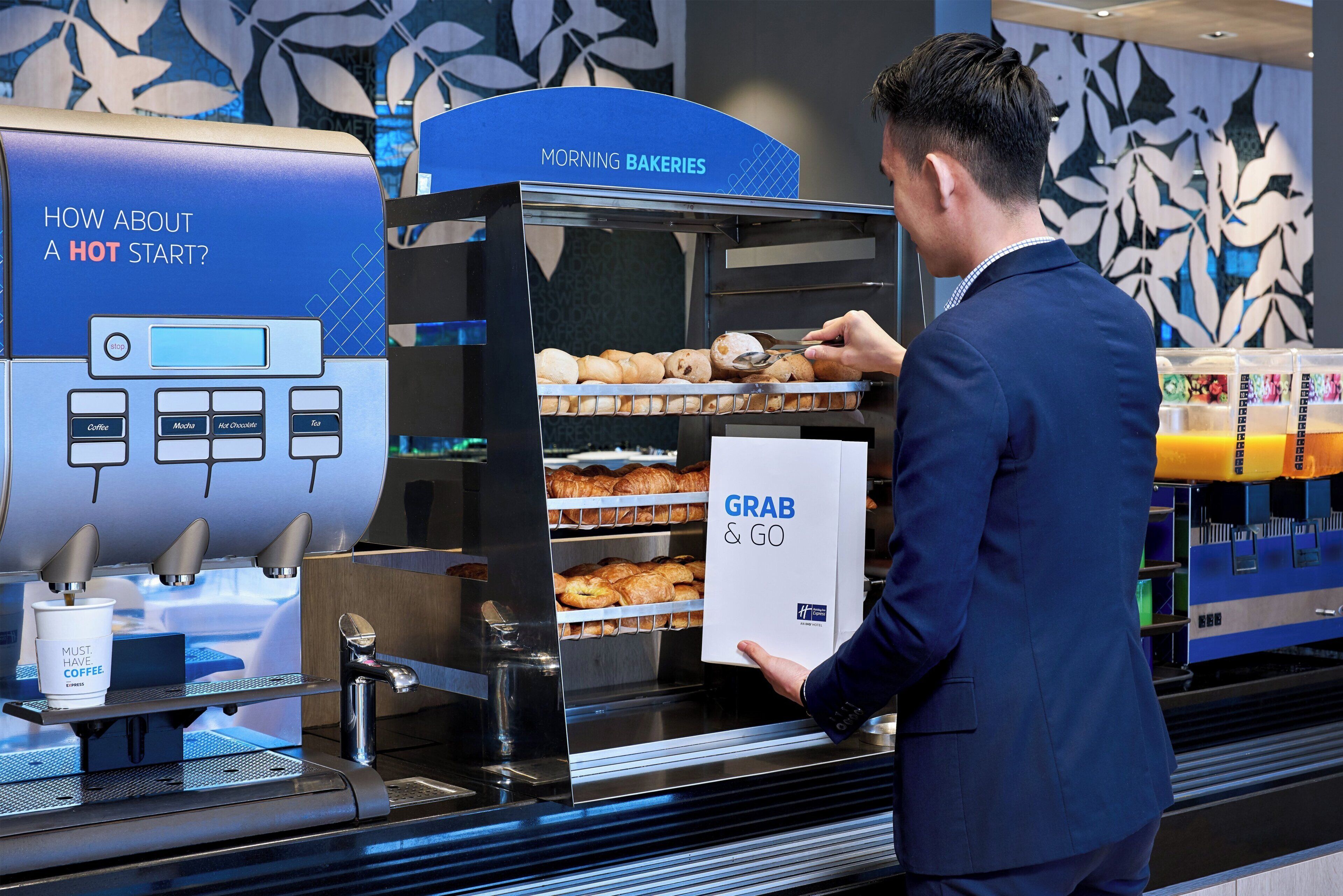 Pessoa de terno azul segurando um letreiro 'GRAB & GO' próximo a uma vitrine de padaria com diversos produtos de panificação.