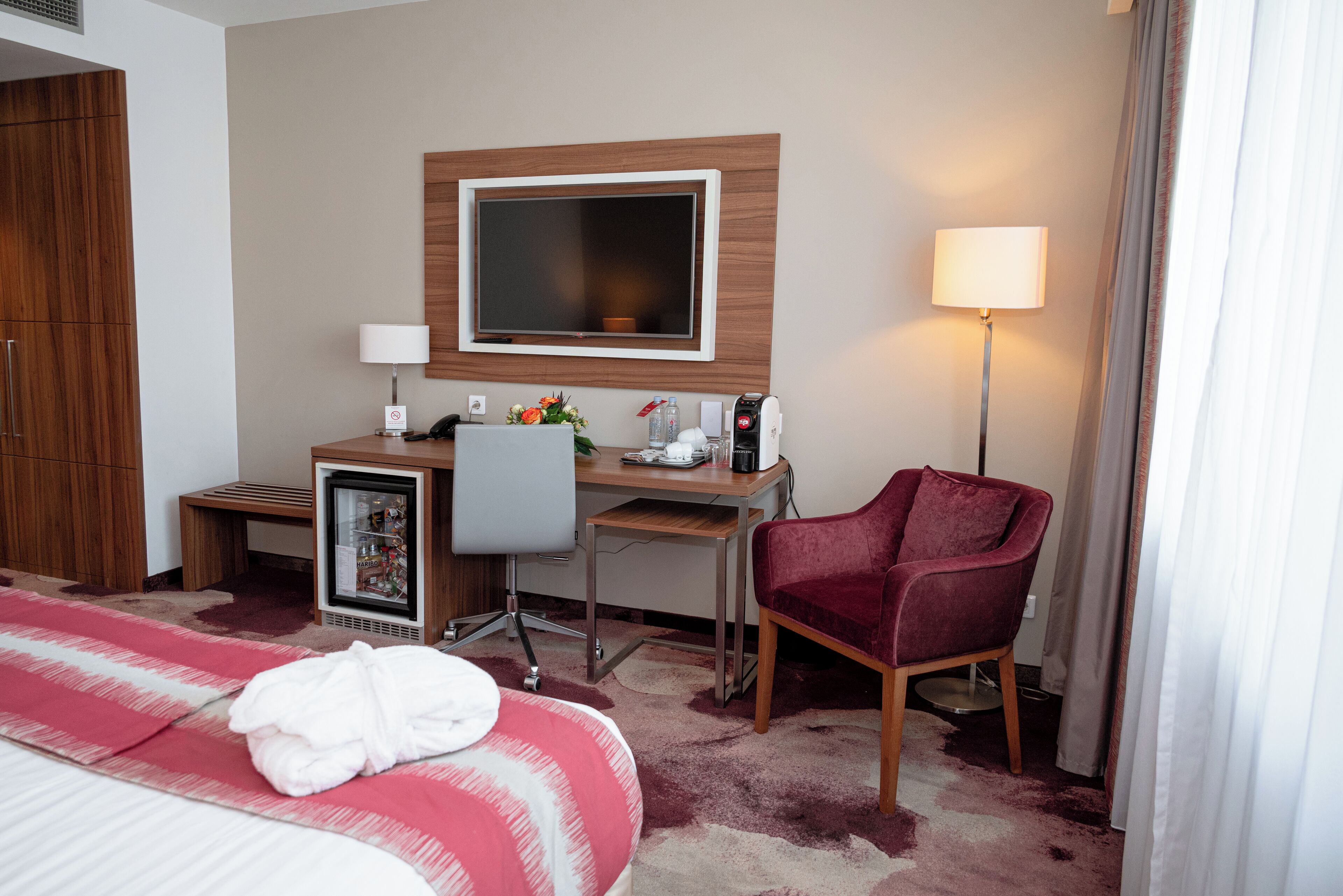 Quarto de hotel com cama, secretária com computador, cadeira, televisão e luminária.