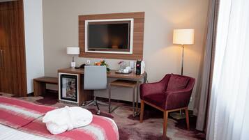 Quarto de hotel com cama, secretária com computador, cadeira, televisão e luminária.