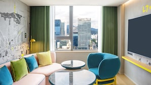 Quarto de hotel moderno com vista para a cidade, um sofá, duas cadeiras, uma mesinha e uma televisão grande.