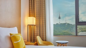 Quarto de hotel com cama, uma cadeira, uma mesinha e uma luminária. Há uma janela com vista para uma torre e vegetação.