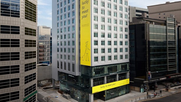 Um hotel moderno com uma placa amarela chamativa na lateral, cercado por outros edifícios.