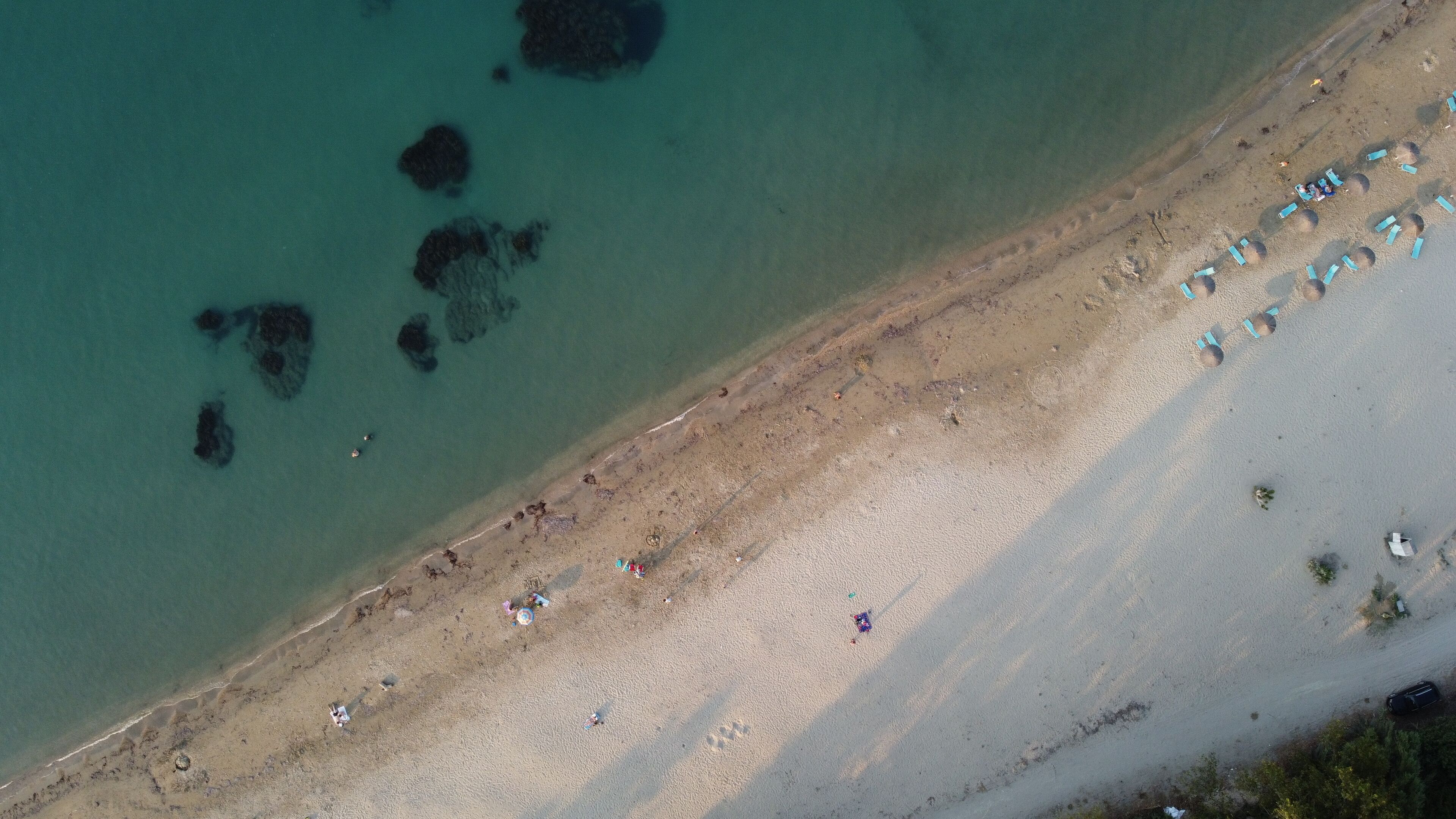 Vista aérea de uma praia com águas turquesa cristalinas, afloramentos rochosos e uma faixa de areia com guarda-sóis e cadeiras de praia.