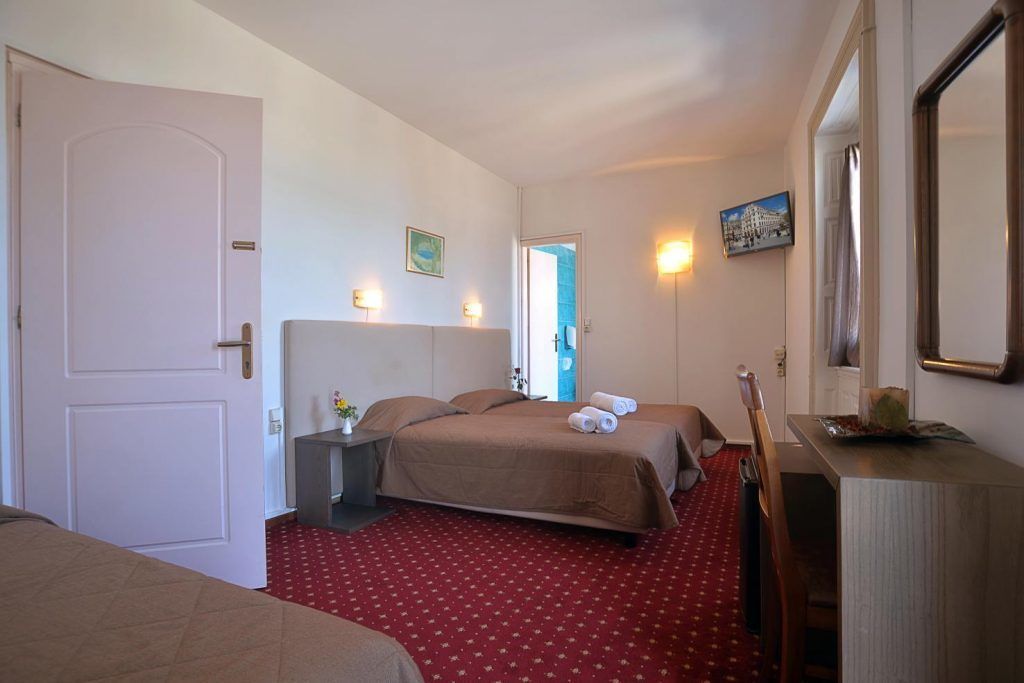 Quarto de hotel com duas camas, uma escrivaninha, uma cadeira, um espelho e piso coberto por um tapete vermelho.